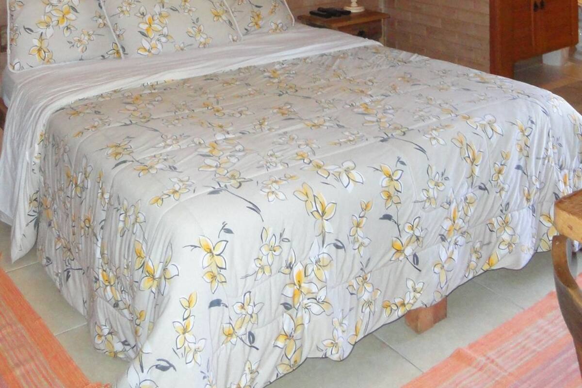Quarto com uma cama grande, cabeceira de madeira, mesa de cabeceira, abajur e parede de tijolos com duas pinturas florais.