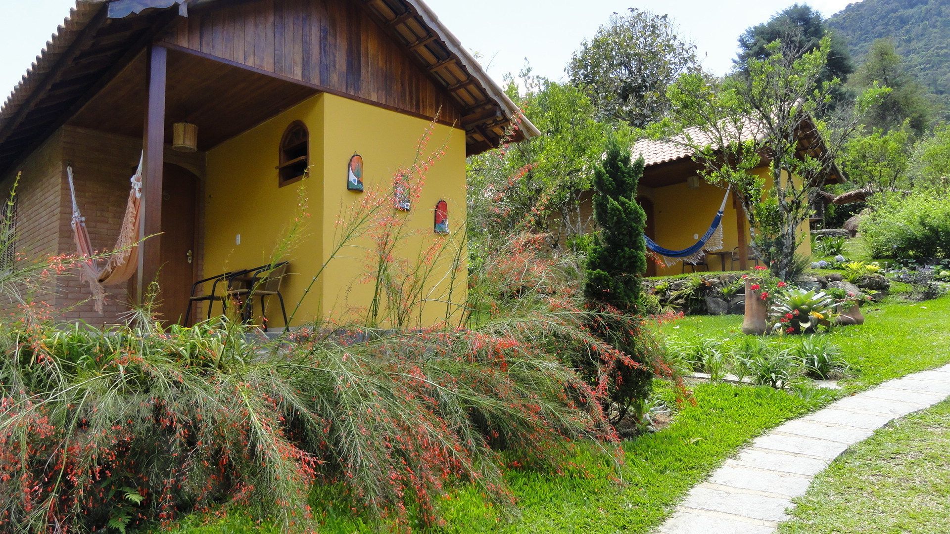 Um edifício amarelo com varanda de madeira, rodeado por vegetação exuberante e um caminho de pedra.