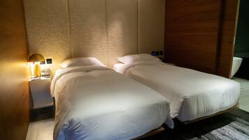 Quarto de hotel com duas camas de solteiro, uma mesa de cabeceira com abajur e um guarda-roupa de madeira.
