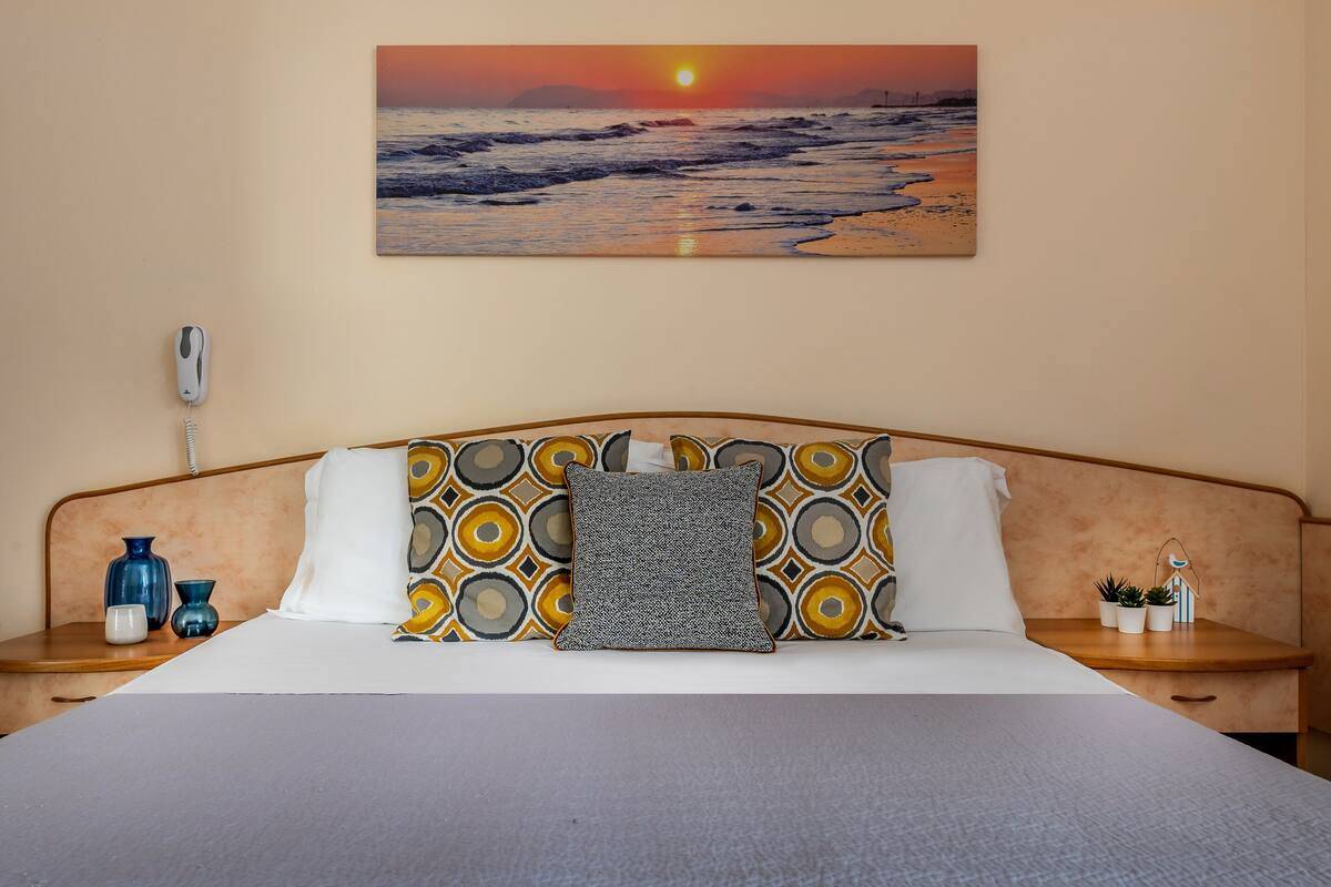 Uma cama bem arrumada com almofadas estampadas, uma mesa de cabeceira com um vaso e um telefone, e um quadro com um pôr do sol sobre o oceano na parede.