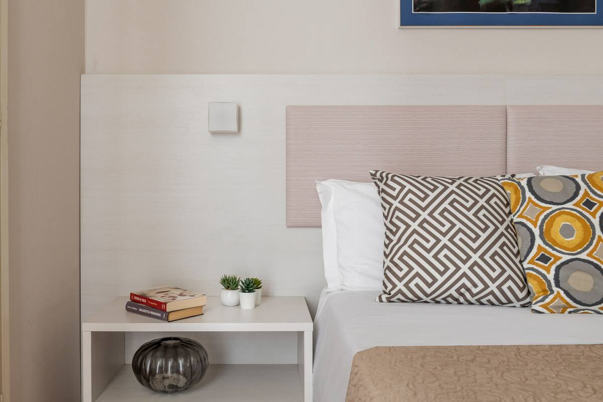 Quarto bem arrumado com uma cama, mesa de cabeceira e um pequeno estante com livros e plantas.