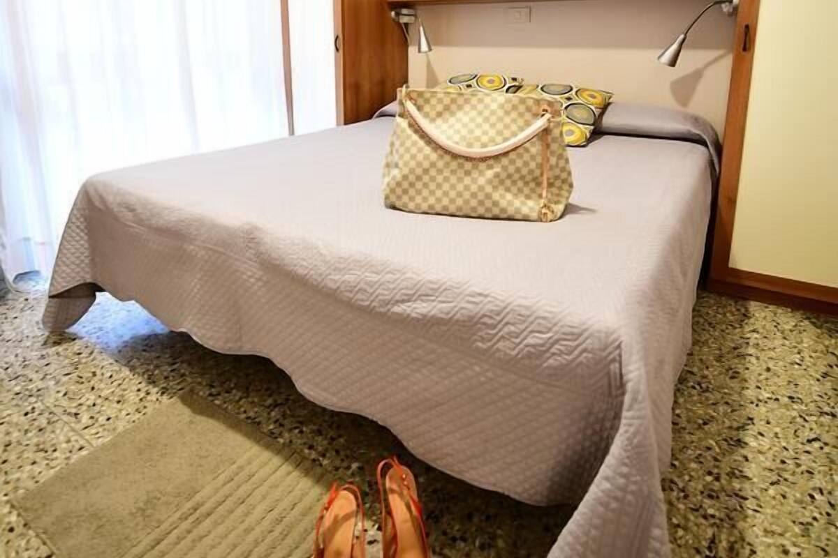Quarto de hotel com uma cama, um guarda-roupa de madeira, uma bolsa quadriculada e sapatos vermelhos no chão.