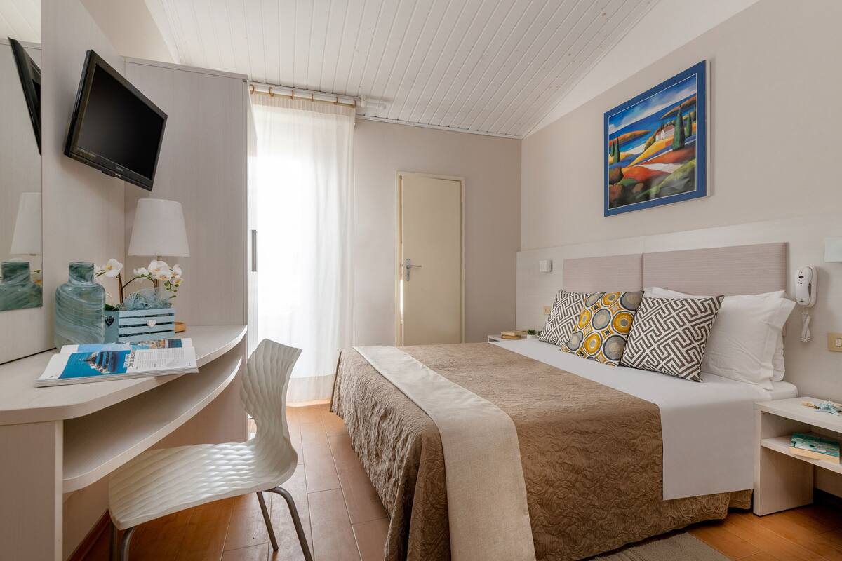 Quarto de hotel com cama, escrivaninha, cadeira, televisão e um quadro na parede.