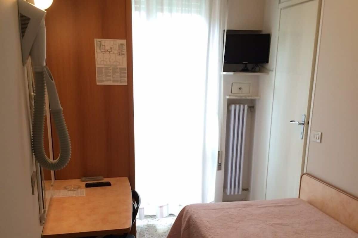 Quarto de hotel compacto com cama, escrivaninha, cadeira, mesinha, televisão e janela com cortinas.