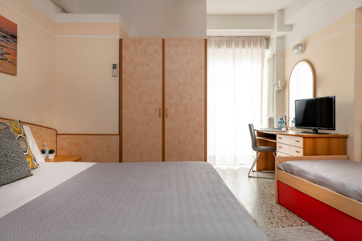 Quarto de hotel com cama, escrivaninha com televisão, guarda-roupa e janela com cortinas.