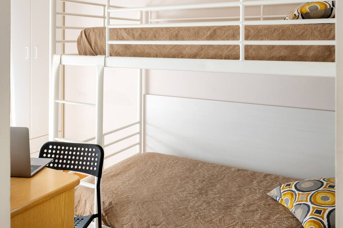 Uma cama de beliche com um laptop em uma escrivaninha, uma cadeira e um travesseiro estampado.