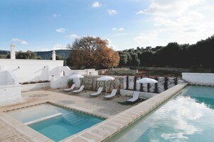 Uma área de piscina com espreguiçadeiras e guarda-sóis, cercada por paredes brancas e um muro de pedra com vegetação.