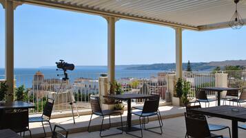 Uma área de estar coberta ao ar livre, com mesas e cadeiras, oferecendo vista panorâmica para o mar e edifícios ao longe.