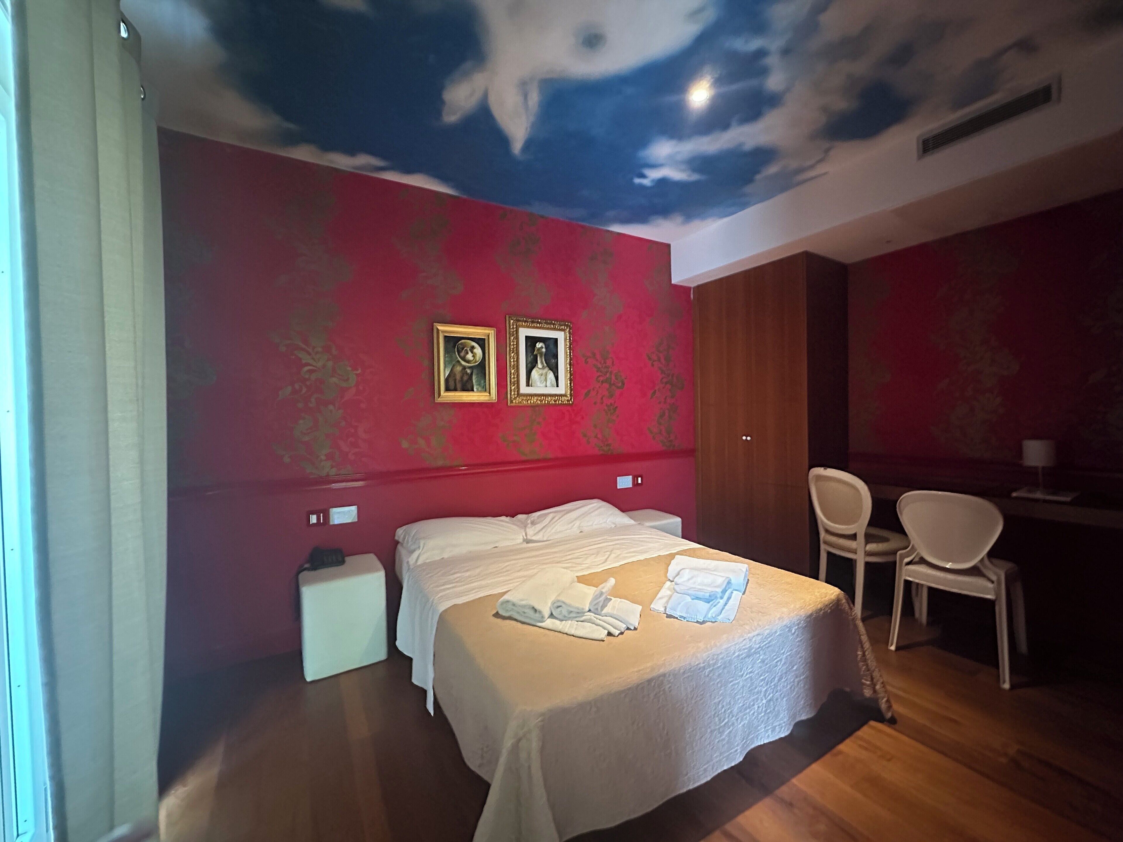 Quarto de hotel com cama, duas cadeiras, escrivaninha e um mural no teto.