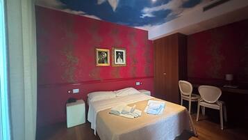 Quarto de hotel com cama, duas cadeiras, escrivaninha e um mural no teto.