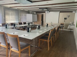 Uma sala de conferências com mesas compridas, cadeiras e uma tela de projeção.