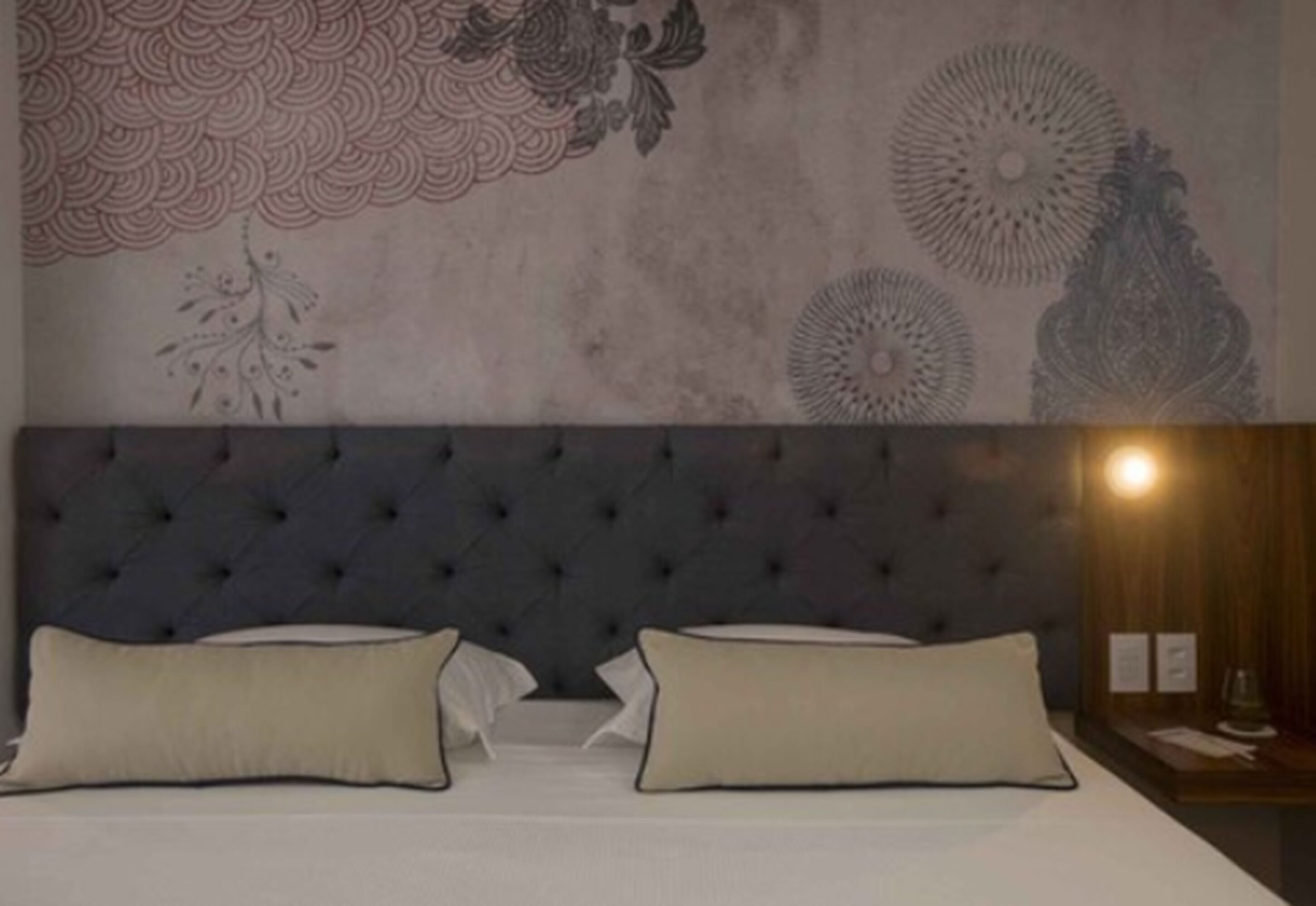Quarto de hotel com cabeceira estofada, duas almofadas e parede decorativa com padrões circulares.