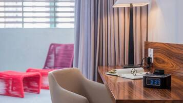 Quarto de hotel com escrivaninha de madeira, relógio digital, luminária e janela com persianas.