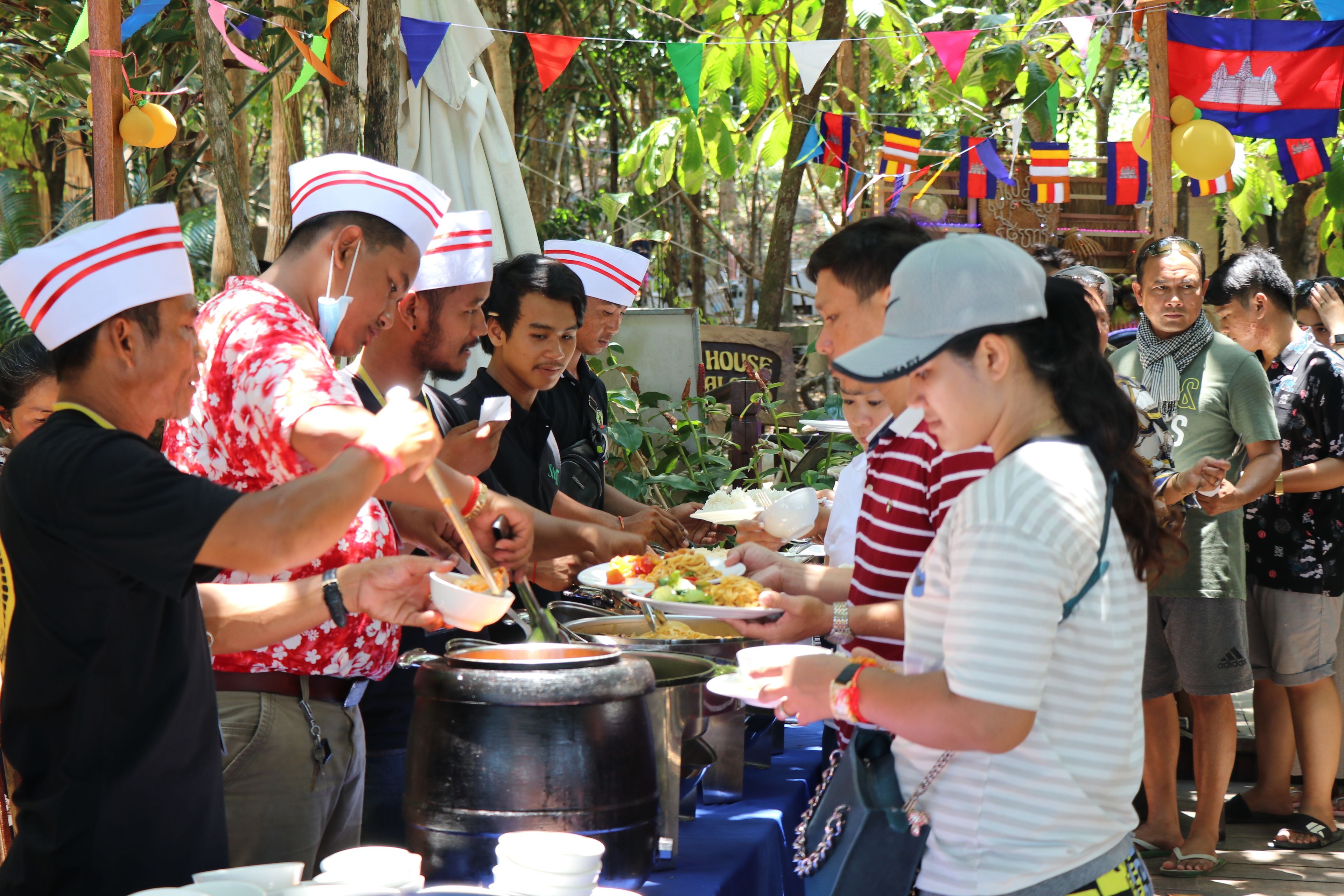Um grupo de pessoas servindo comida em um evento ao ar livre, com uma mesa e recipientes para alimentos.