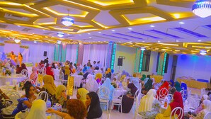 Uma sala de banquetes com mesas redondas, cadeiras brancas e convidados sentados.