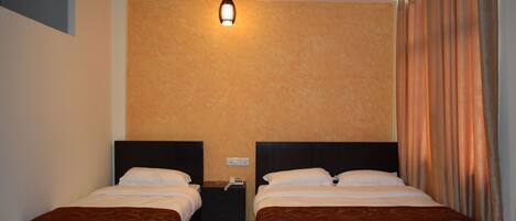 Quarto de hotel com duas camas, uma mesa de cabeceira, uma luminária na parede e uma janela com cortinas.