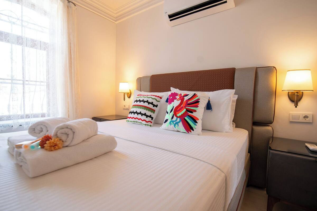 Quarto de hotel com cama bem arrumada, almofadas decorativas, abajur e mesa de cabeceira.