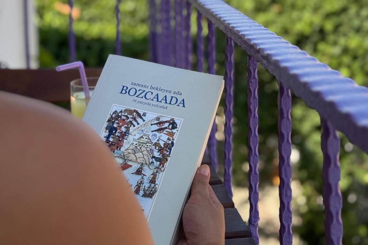 Uma pessoa segurando um livro intitulado 'BOZCAADA' numa varanda com guarda-corrimão roxo, com vista para uma paisagem urbana com edifícios e uma torre.