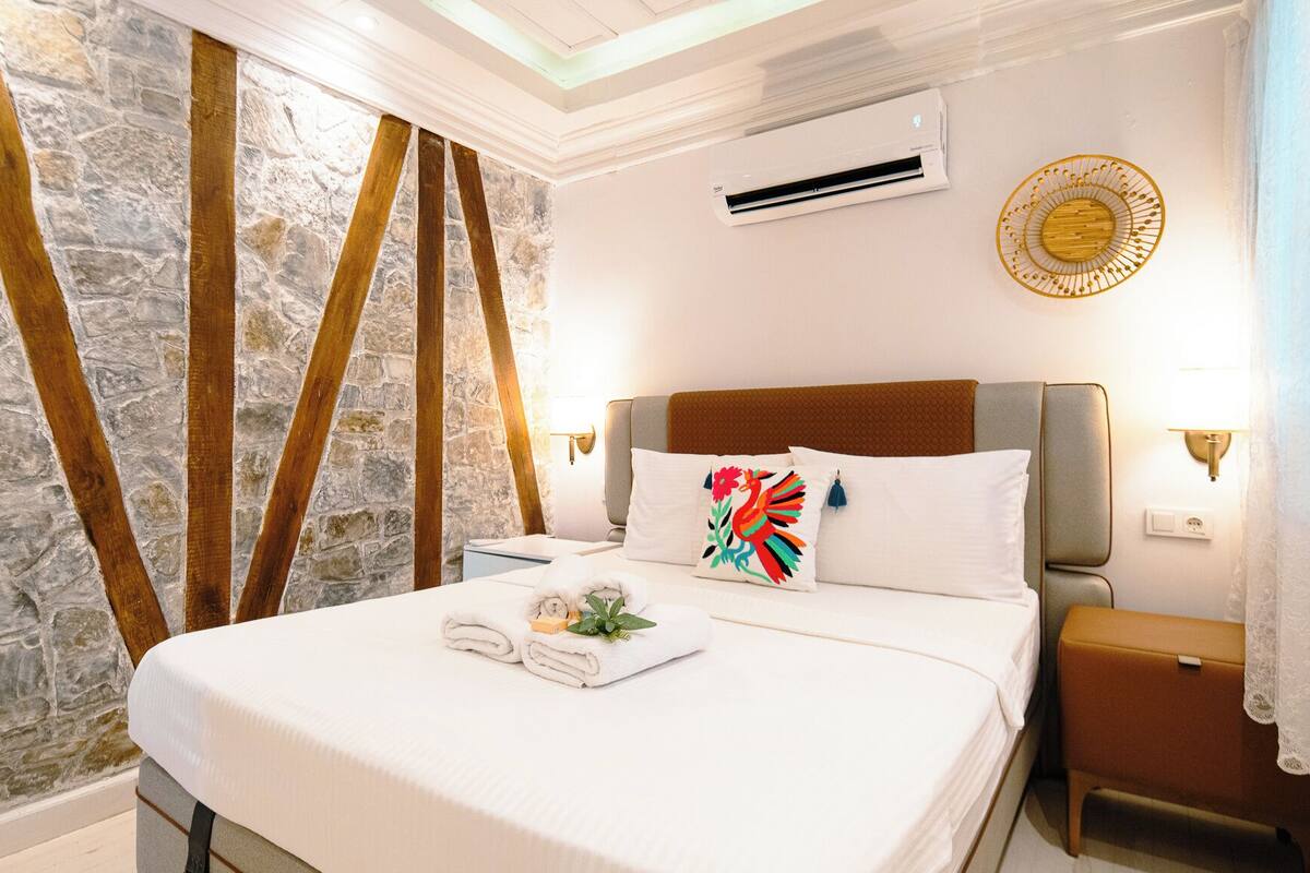 Quarto de hotel com parede de pedra, cama com roupa de cama branca, almofada decorativa, mesa de cabeceira e aparelho de ar condicionado.