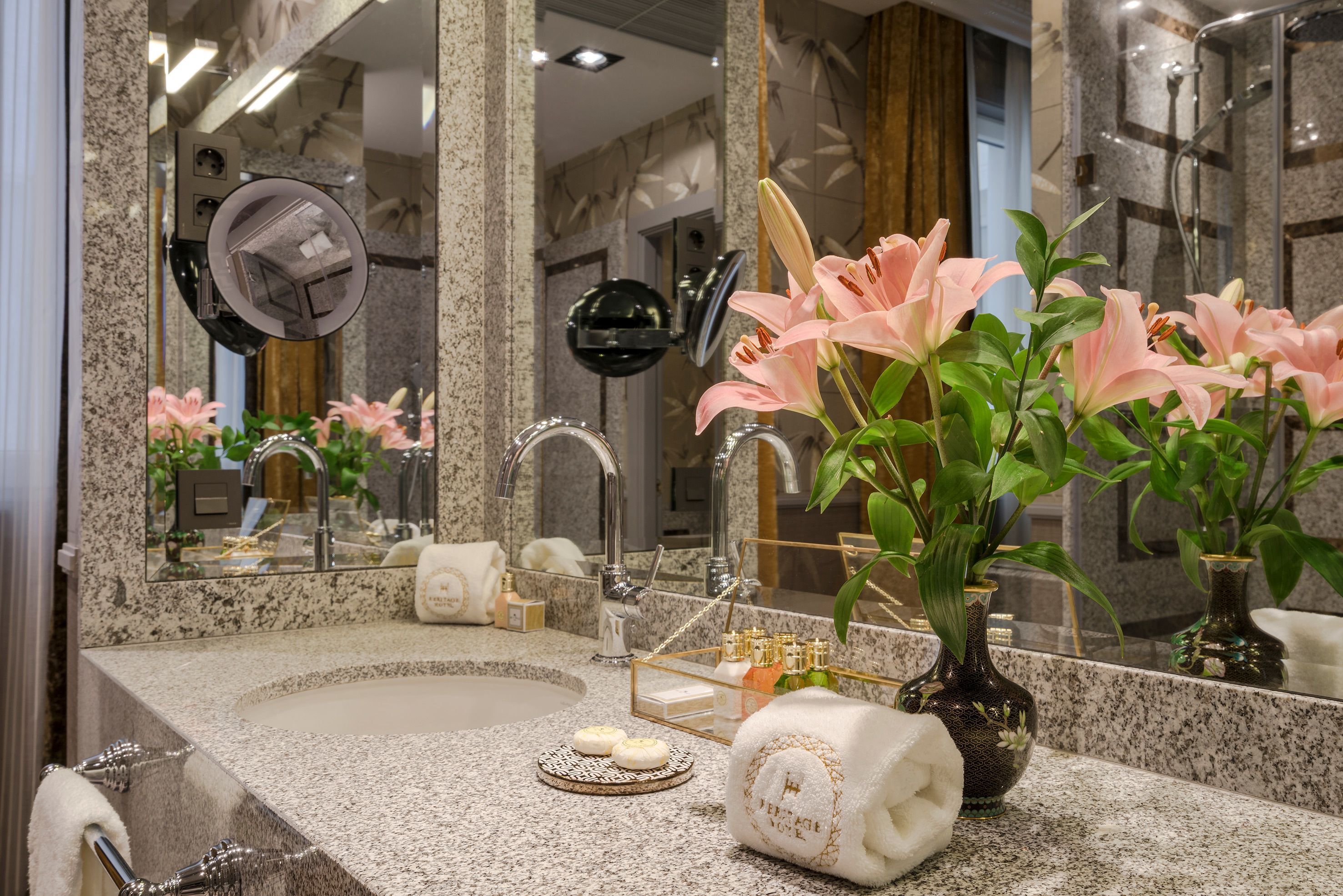 Casa de banho com bancada de granito, lavatório, espelho, toalha e um vaso com flores.