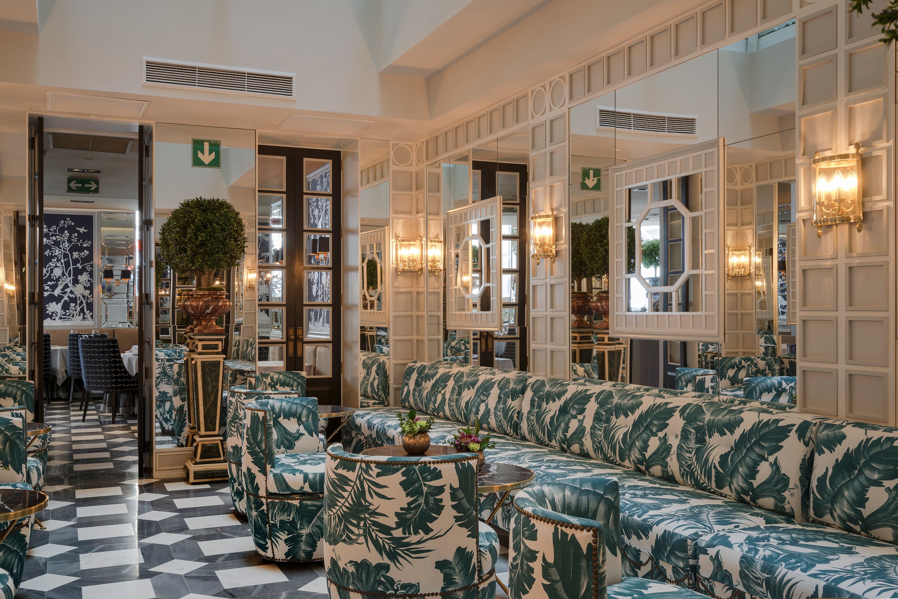 Um saguão de hotel com piso xadrez, um sofá grande com padrão de folhagem tropical e uma planta em vaso.