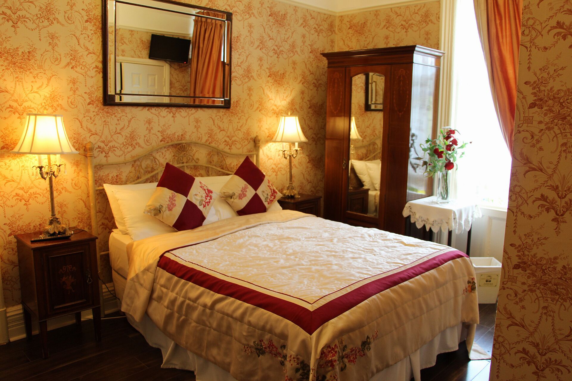 Cama bem arrumada com colcha e almofadas estampadas com padrão floral, duas luminárias de cabeceira, mesa de cabeceira de madeira, um espelho e um vaso com flores sobre uma mesinha.