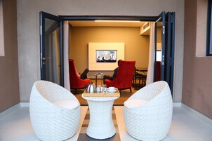 Quarto de hotel moderno com uma grande janela, TV de tela plana e um espaço de estar com duas cadeiras brancas e uma mesa pequena.