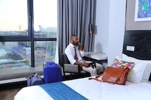 Quarto de hotel com uma cama, uma mesa, uma cadeira, uma janela com vista para um aeroporto e uma mala.