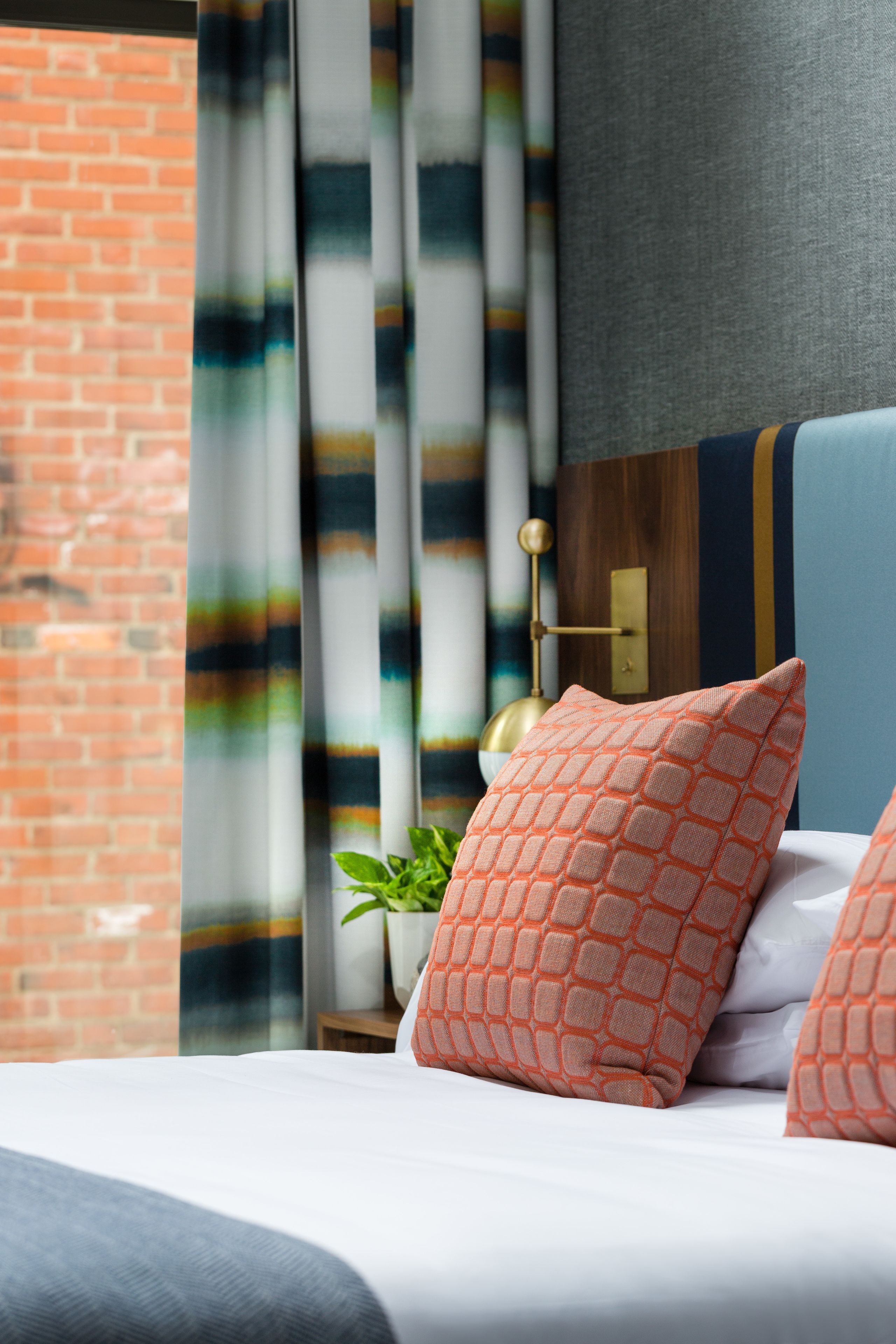 Quarto de hotel com parede de tijolos, cama com roupa de cama branca e mesa de cabeceira com uma planta e um abajur.