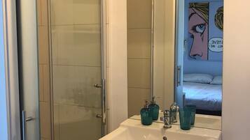 Um banheiro com box de vidro, pia branca com duas garrafas e espelho refletindo uma cama.