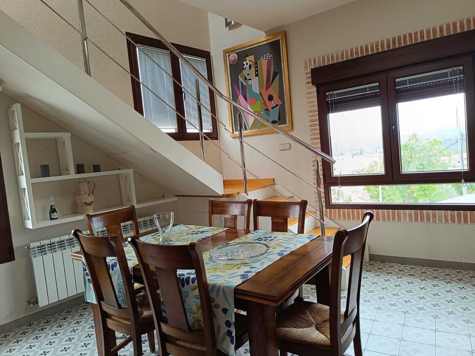 Uma área de refeições com uma mesa e cadeiras de madeira, uma escada, um radiador e uma janela com vista.