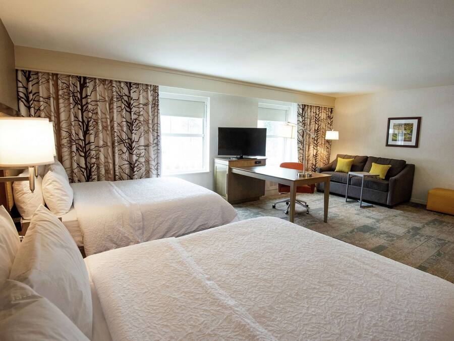 Quarto de hotel com duas camas, uma escrivaninha, televisão, sofá e janela com cortinas.