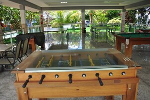 Uma mesa de pebolim em área externa coberta, com uma mesa de bilhar por perto.