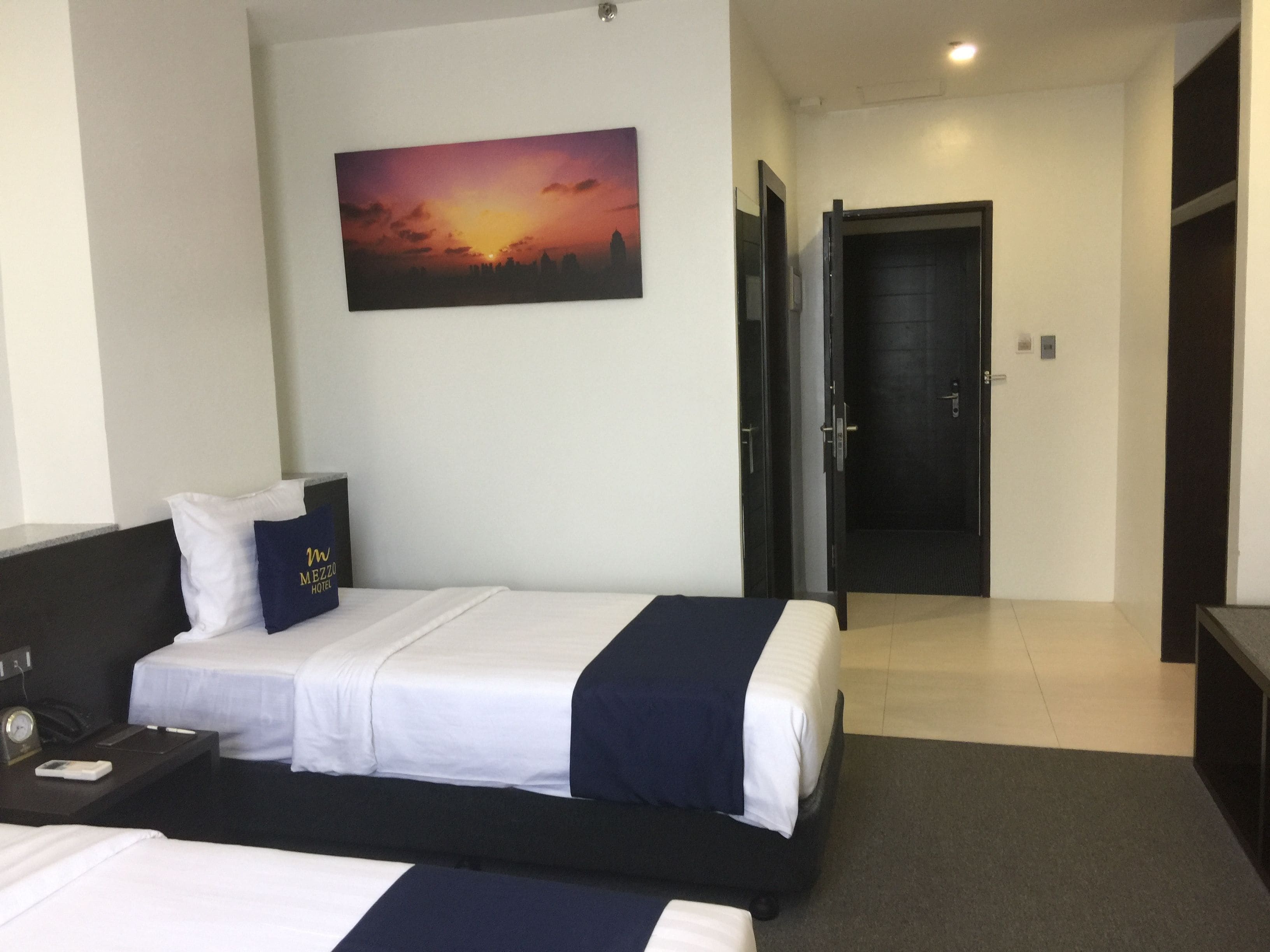 Quarto de hotel com cama, mesa de cabeceira, quadro de um pôr do sol e porta para outro cômodo.