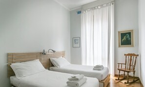 Quarto de hotel com duas camas de solteiro, cabeceira de madeira, luminária de cabeceira, quadro emoldurado, cadeira de balanço e janela com cortinas.