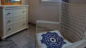 Quarto com uma cadeira de vime branca, penteadeira de madeira com gavetas, janela com vista e teto de madeira.