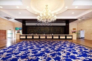 Um hall amplo com um grande tapete azul estampado, um candelabro, um balcão de recepção com tela digital e uma parede com detalhes decorativos.