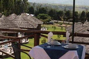 Uma área de refeições com uma mesa posta para dois, com vista para uma paisagem com cabanas de palha e colinas ao longe.