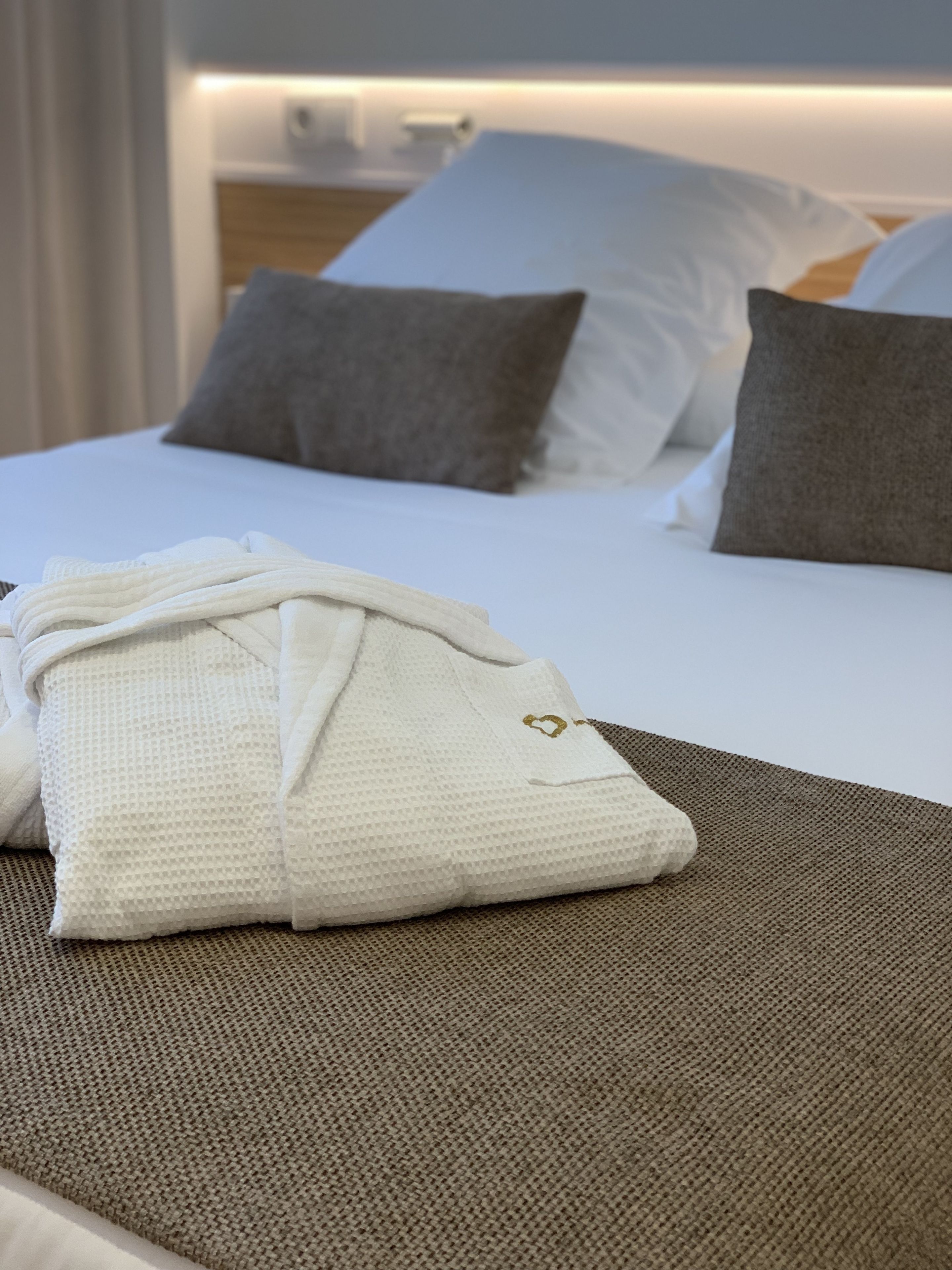 Cama bem arrumada com roupa de cama branca e um roupão branco dobrado sobre um tapete marrom texturizado.