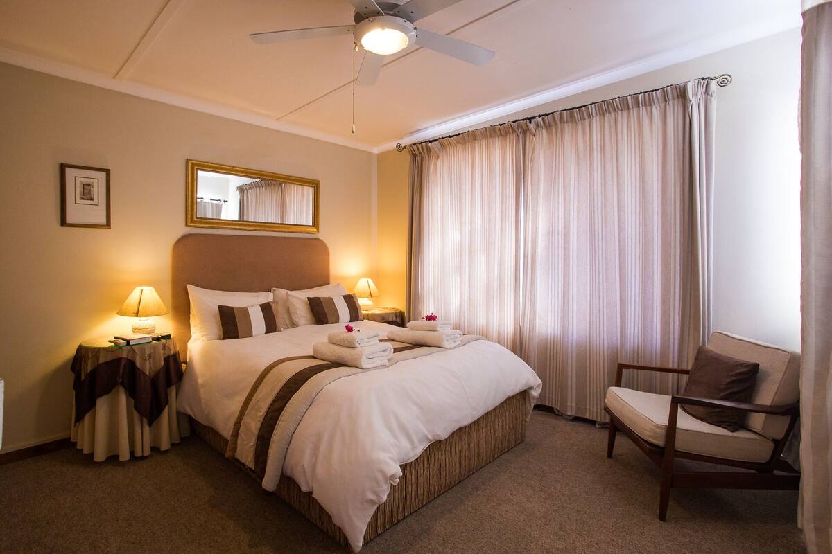 Quarto de hotel com cama, luminárias de cabeceira, uma cadeira, ventoinha no teto e uma janela grande com cortinas.