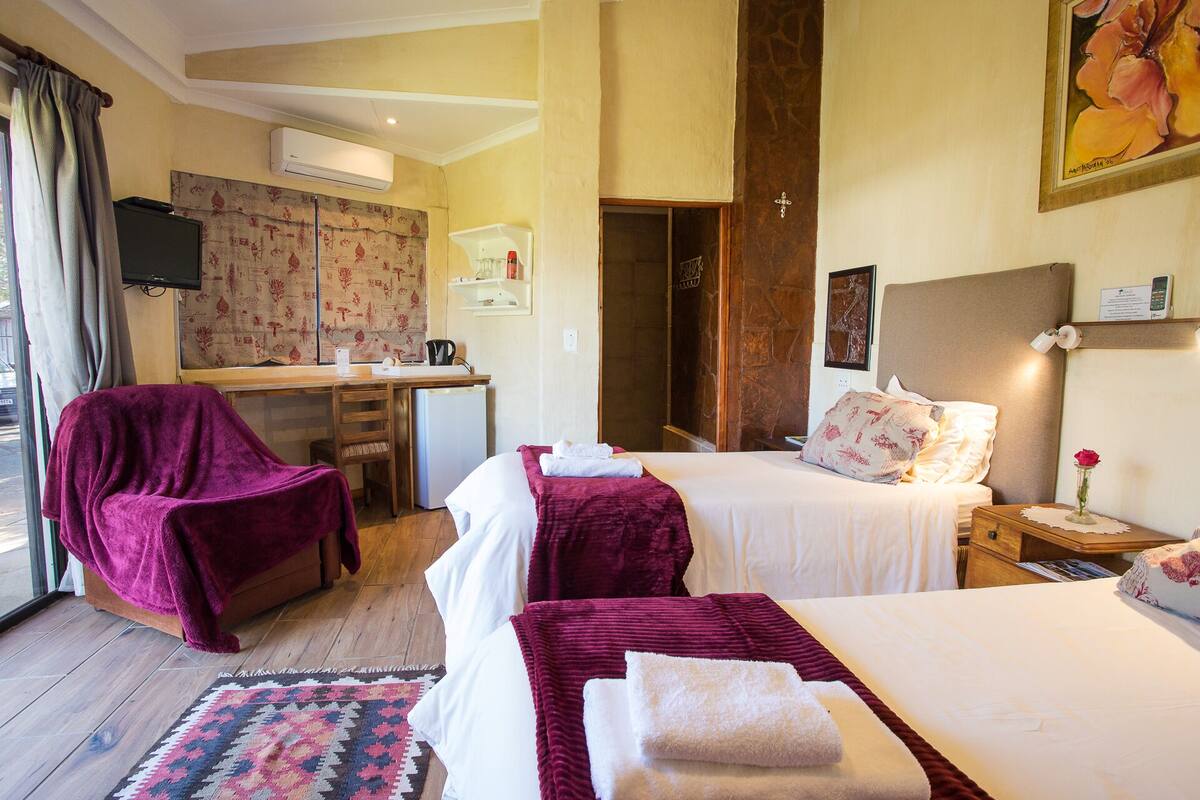 Quarto de hotel com uma cama, duas mesas de cabeceira, uma escrivaninha com uma cadeira, um pequeno frigorífico, uma janela com cortinas e um quadro na parede.