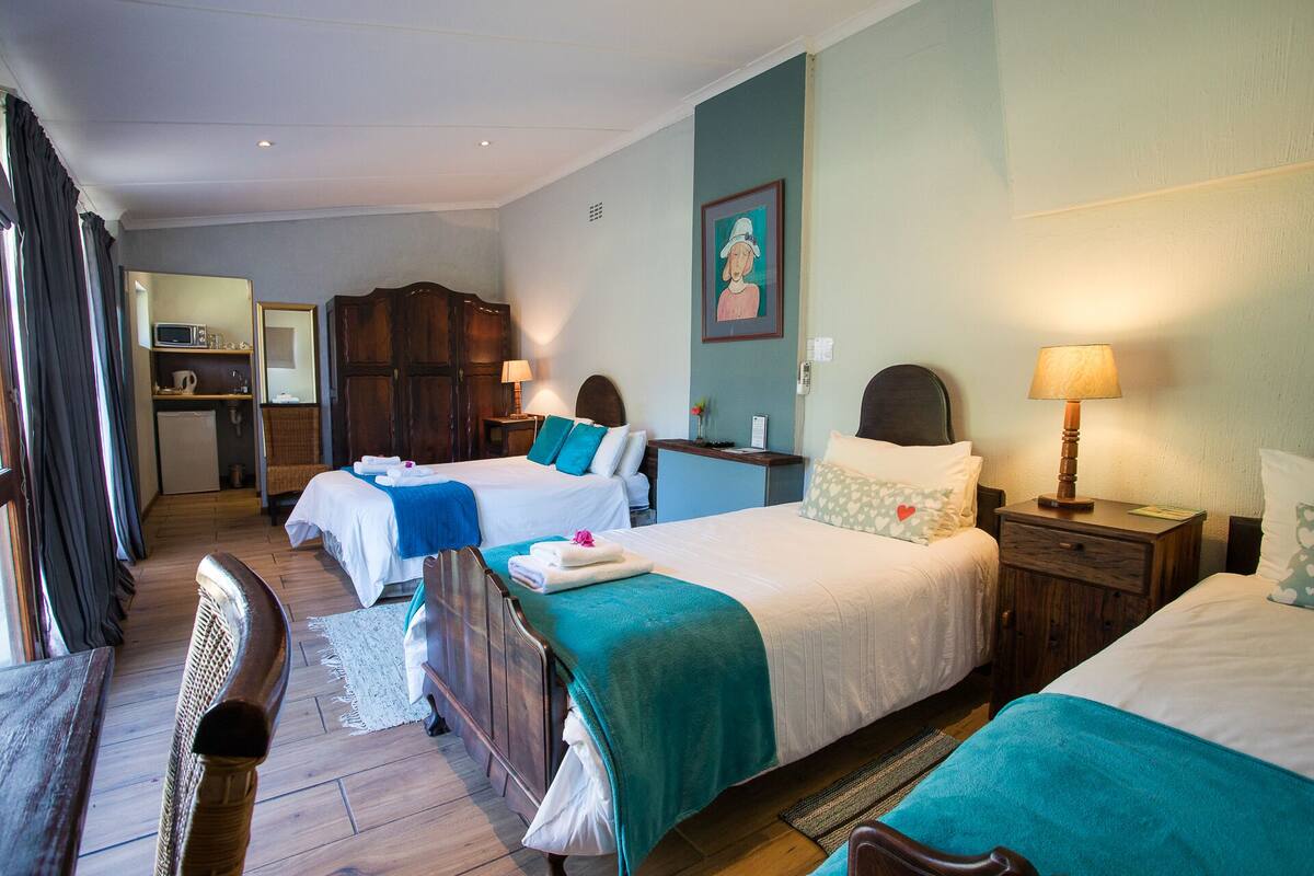 Quarto de hotel com duas camas, uma escrivaninha, uma cadeira, uma kitchenette pequena e um banheiro.