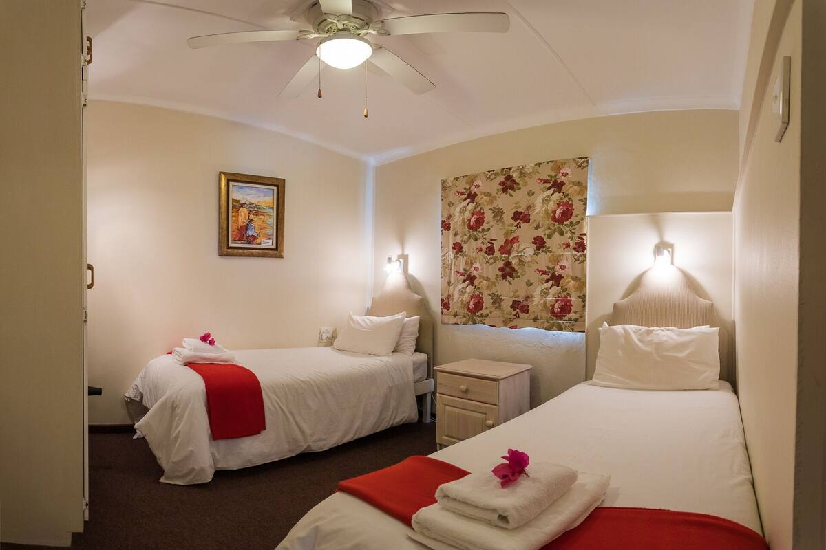 Quarto de hotel com duas camas, ventoinha no teto, um quadro, cortina floral e abajur.