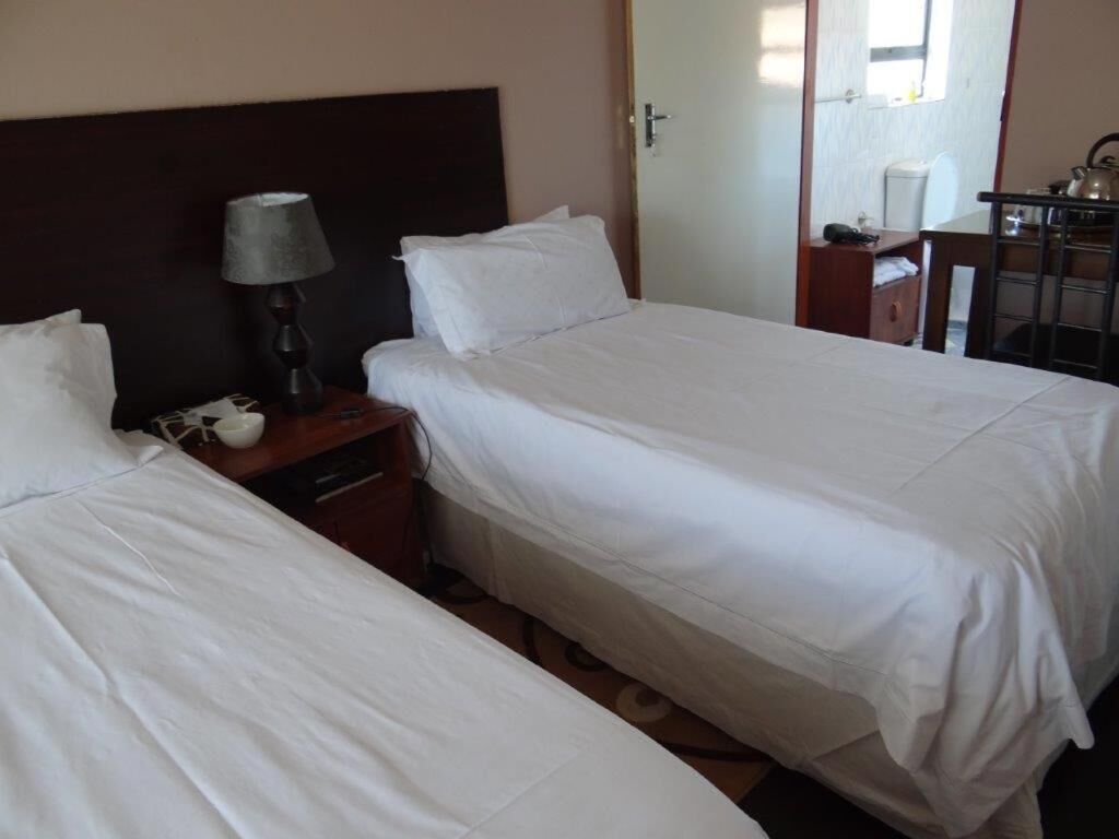 Quarto de hotel com duas camas de solteiro, uma mesa de cabeceira com luminária, uma pequena mesa com chaleira e vista para o exterior através de uma porta.