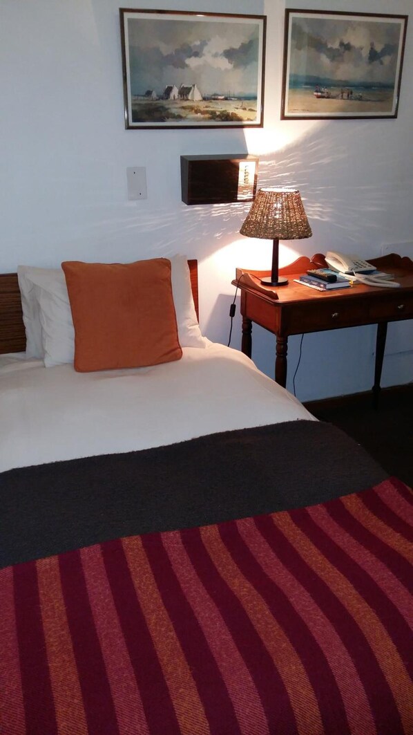Quarto de hotel com uma cama, mesa de cabeceira com abajur, telefone e duas fotos emolduradas na parede.