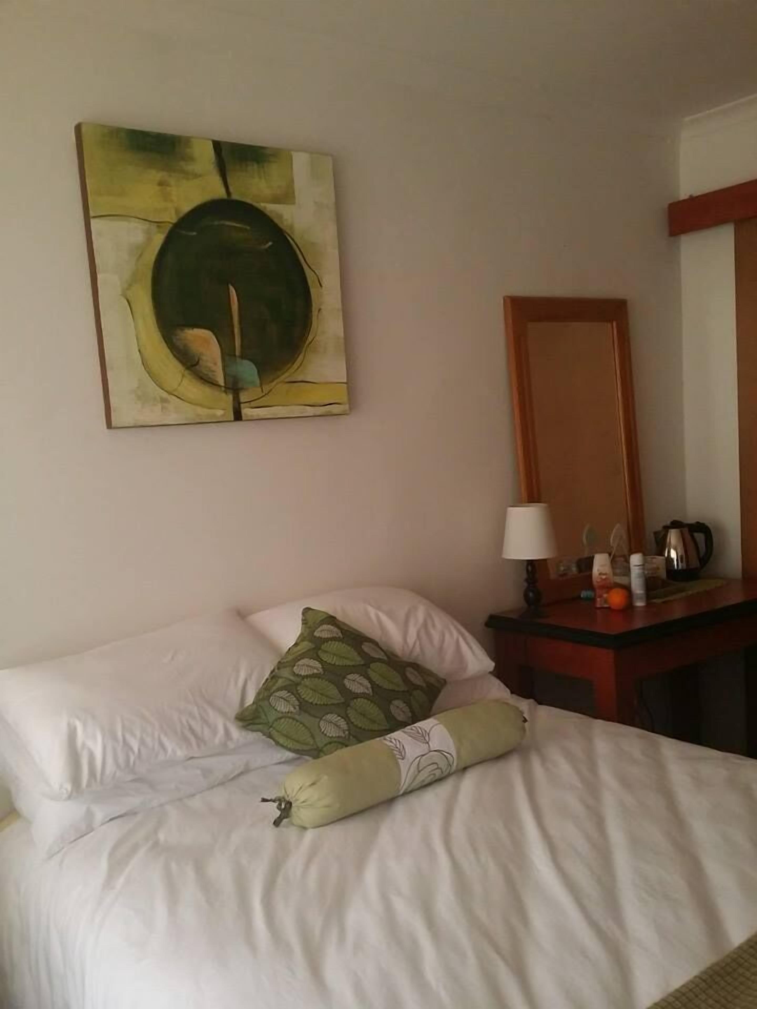 Quarto de hotel com cama, mesa de cabeceira com luminária, cafeteira e um quadro na parede.