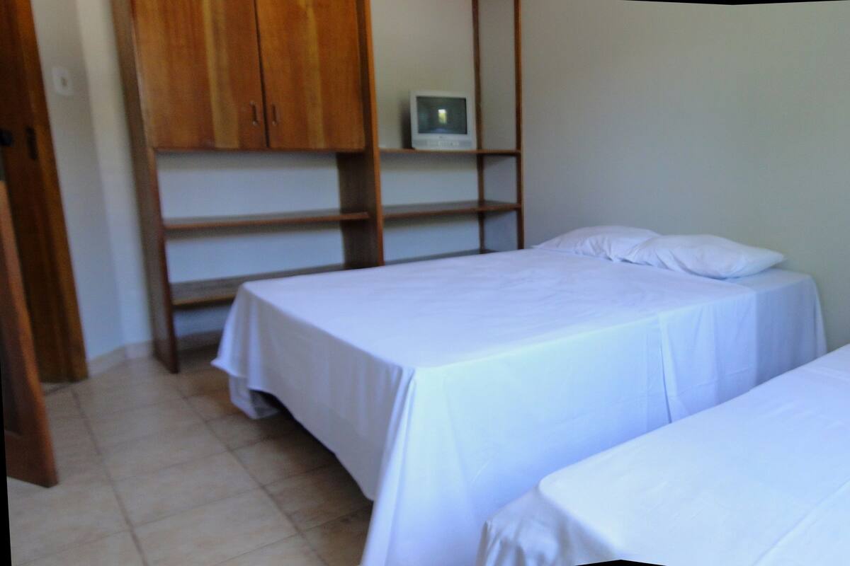 Quarto com duas camas de solteiro, um guarda-roupa de madeira e uma pequena televisão em uma prateleira.