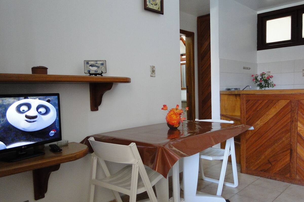 Uma sala de jantar com uma mesa de madeira, cadeiras brancas, uma TV sobre um móvel e uma pequena estante com um objeto decorativo.