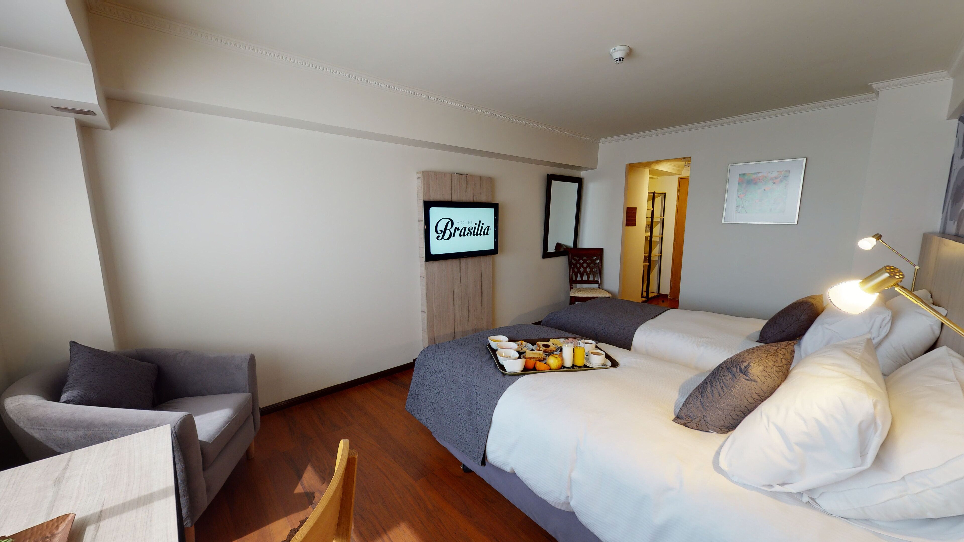 Quarto de hotel com cama, escrivaninha, cadeira e televisão exibindo "HOTEL Brasilia".