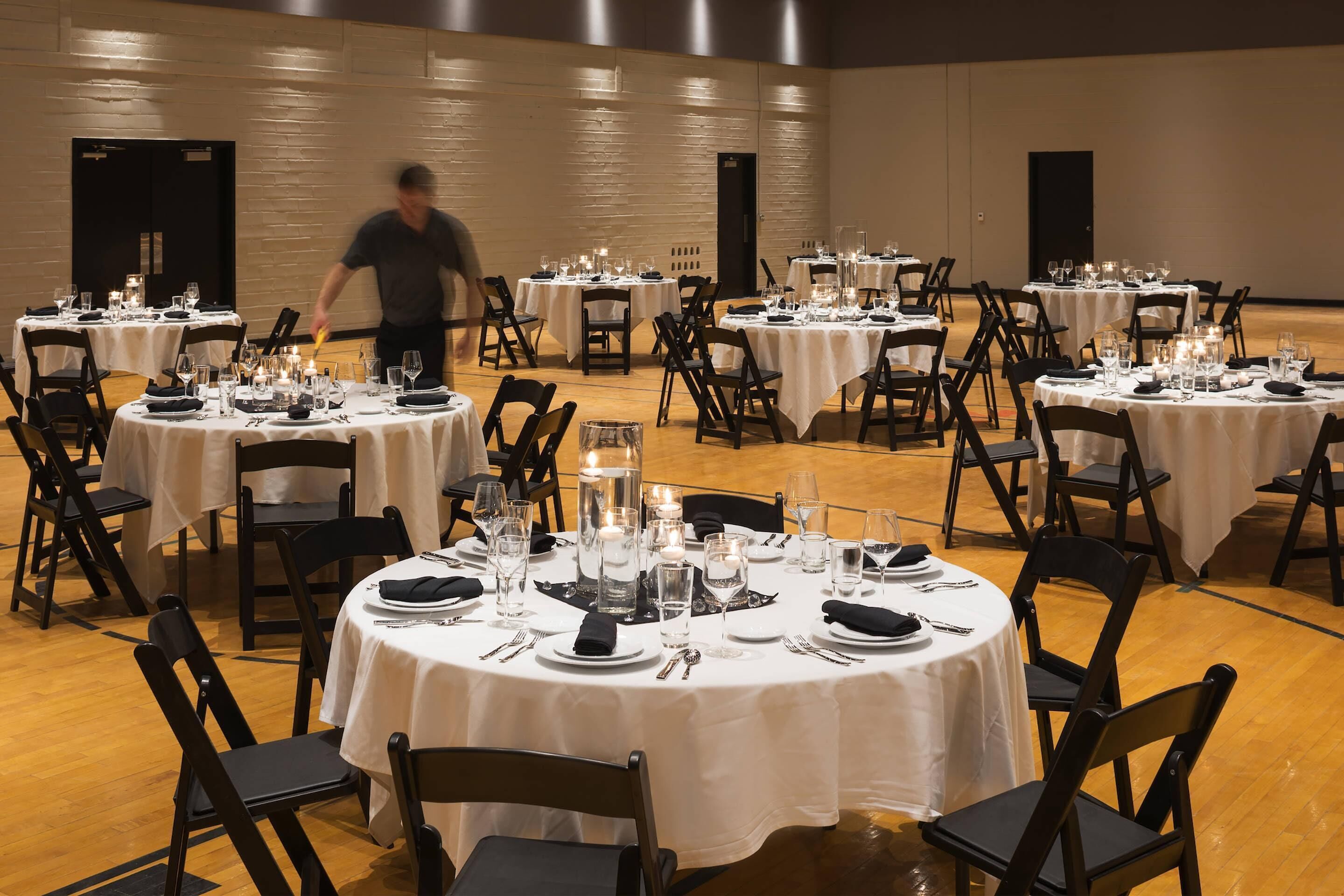 Uma sala de banquetes com mesas redondas preparadas para refeições, cada uma com toalhas brancas, cadeiras pretas e copos.